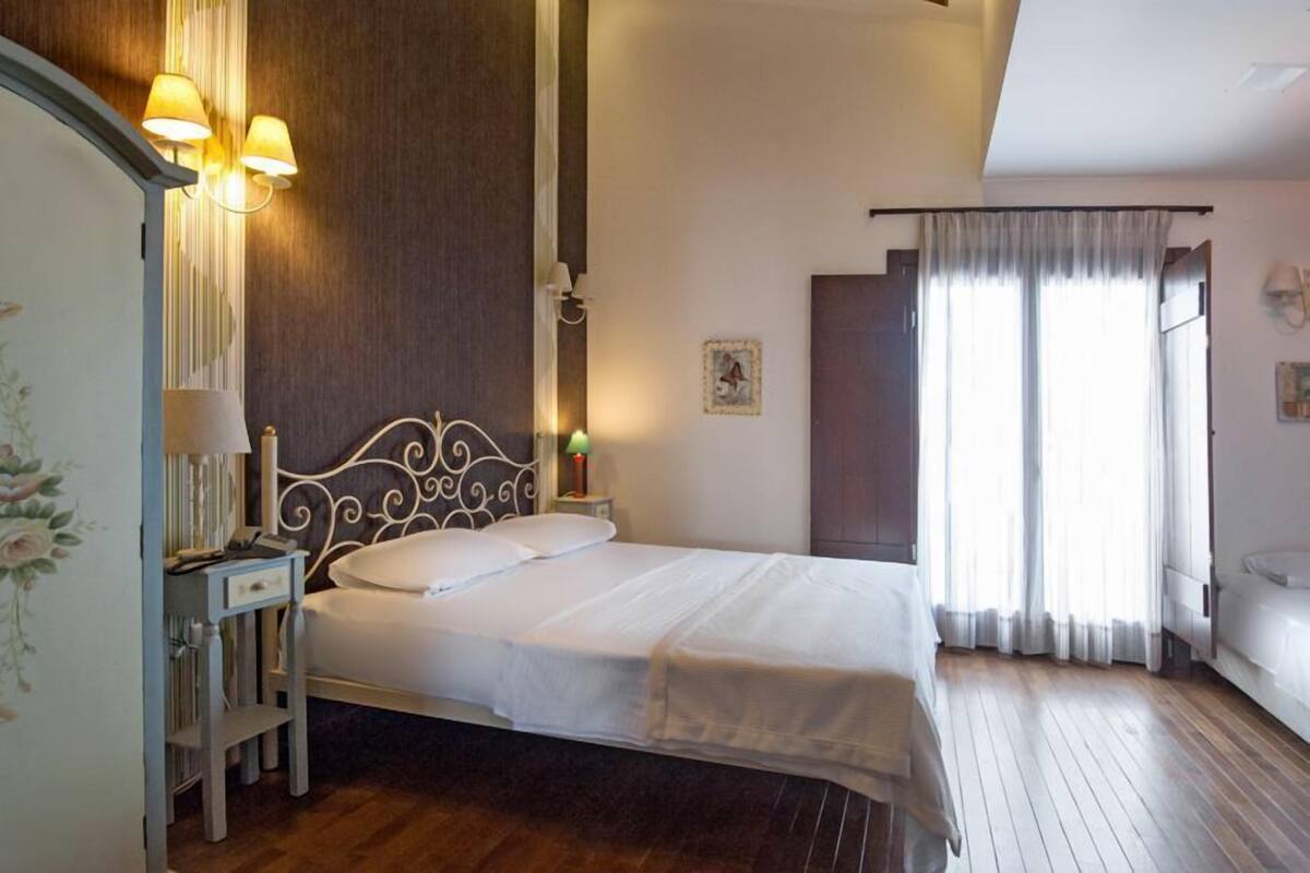 Quarto com cama, abajures, mesa de cabeceira, piso de madeira e janela com cortinas.