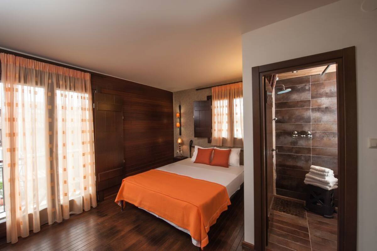 Quarto de hotel com cama, piso de madeira, banheiro com chuveiro e cortinas.