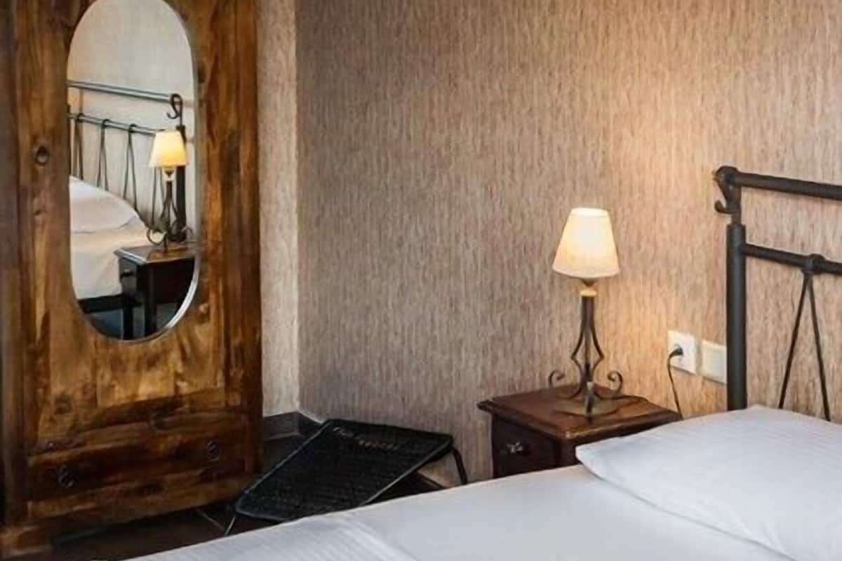 Quarto de hotel com guarda-roupa de madeira, cama com roupa de cama branca, mesa de cabeceira com abajur e luminária na parede.
