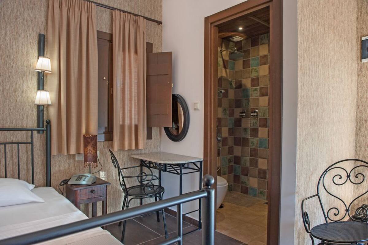 Quarto de hotel com cama, escrivaninha com cadeira, telefone e banheiro com chuveiro e vaso sanitário.