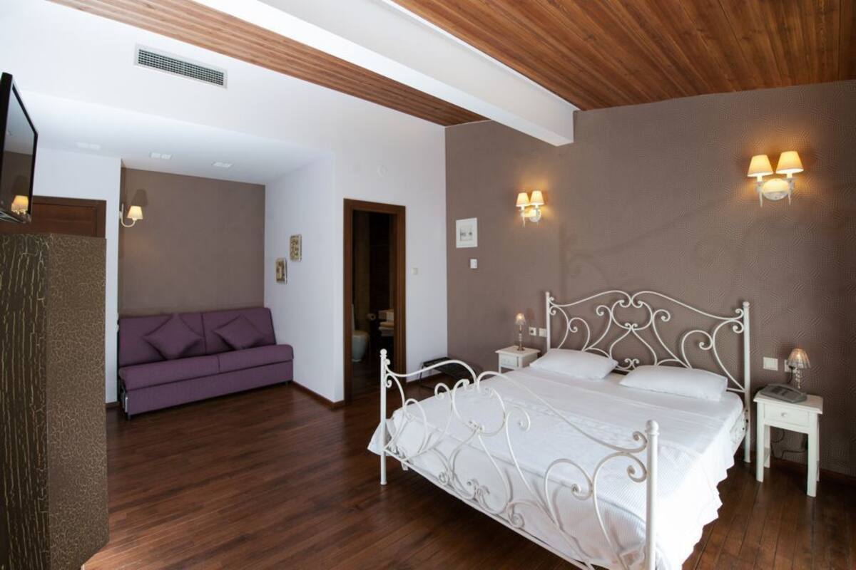 Quarto com estrutura de cama de metal branco, sofá roxo e duas luminárias de cabeceira.