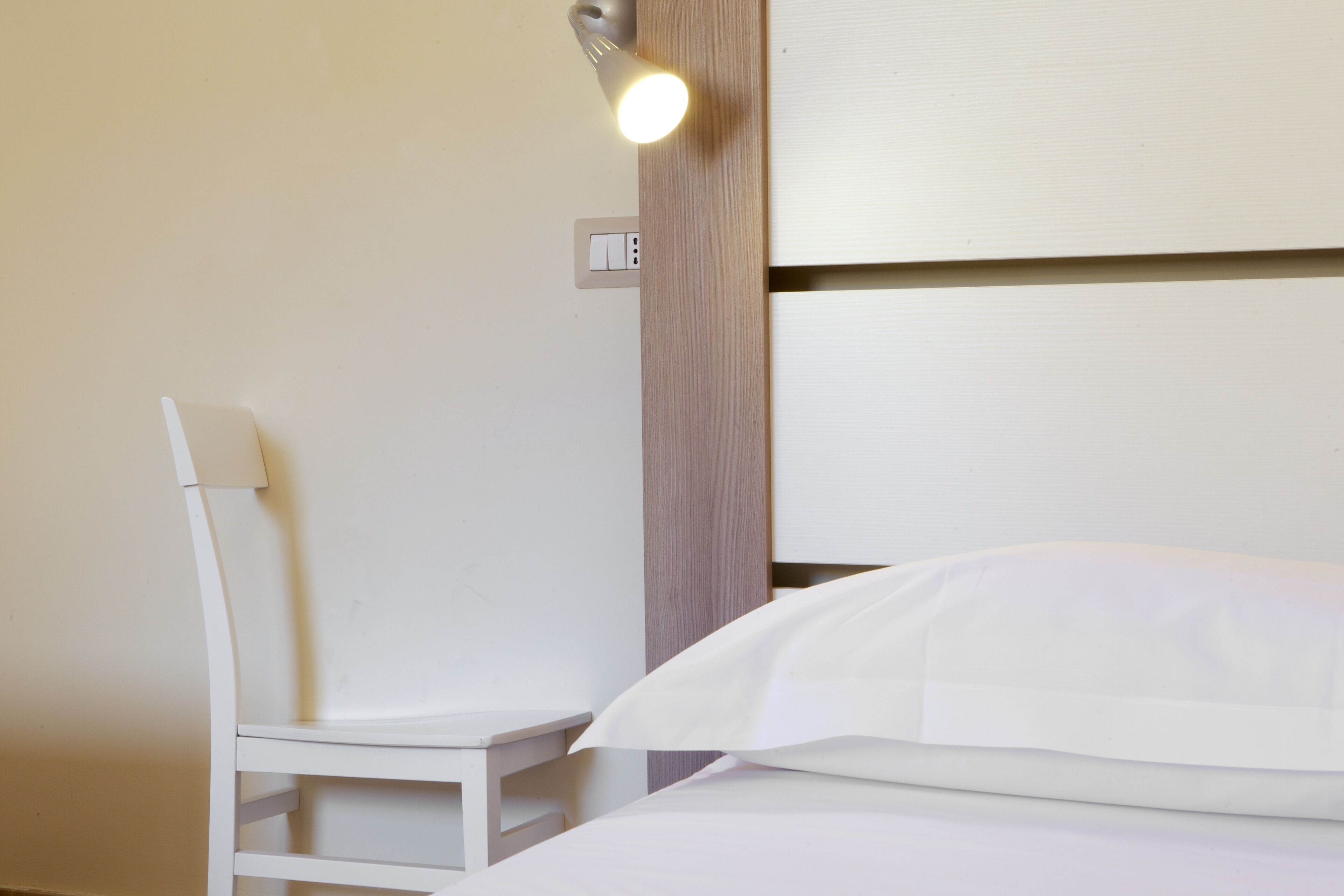 Cama bem arrumada com lençóis brancos, mesa de cabeceira e luminária fixa na parede.