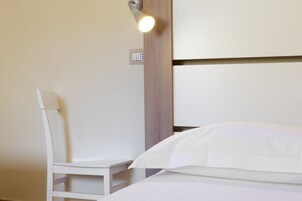 Cama bem arrumada com lençóis brancos, mesa de cabeceira e luminária fixa na parede.