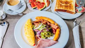 Mesa de café da manhã com ovos, bacon, presunto e legumes, uma xícara de café, uma fatia de melancia e torradas, além de um potinho de iogurte.