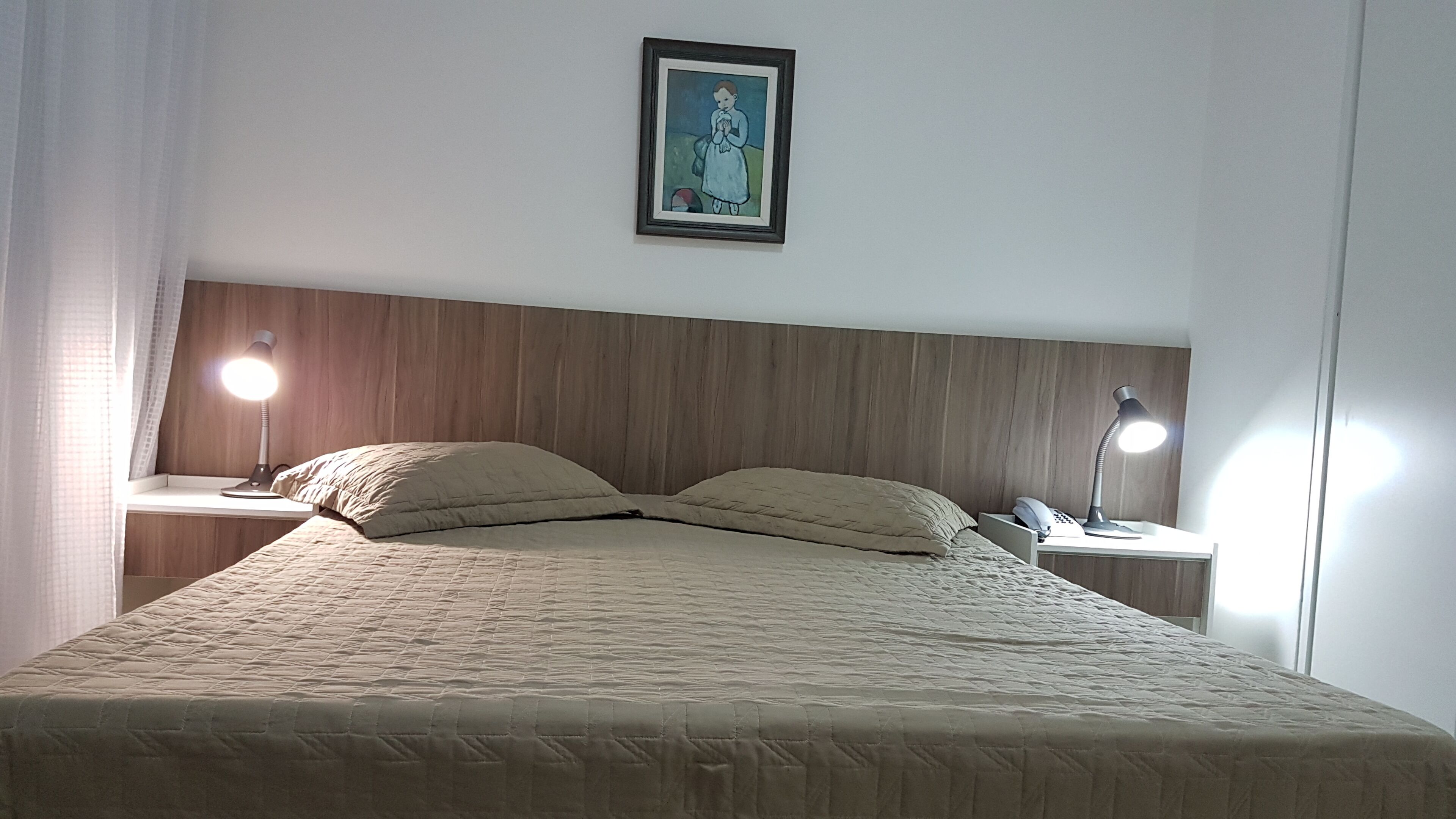Cama bem arrumada com manta acolchoada, duas luminárias de cabeceira e um quadro na parede.