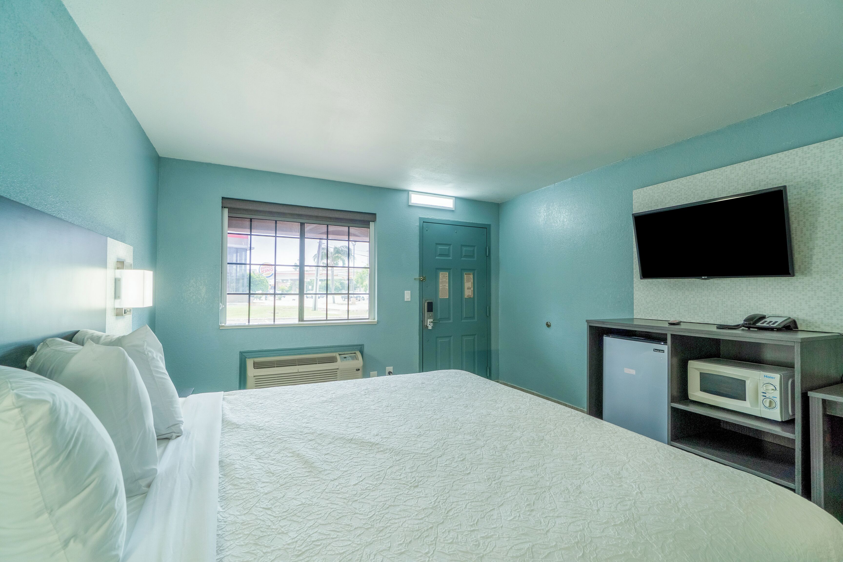 Quarto de hotel com uma cama, uma escrivaninha com micro-ondas, televisão e janela com persianas.