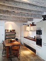 Uma cozinha com teto de madeira r ústica, armários brancos, uma mesa de jantar de madeira com cadeiras e um forno embutido.