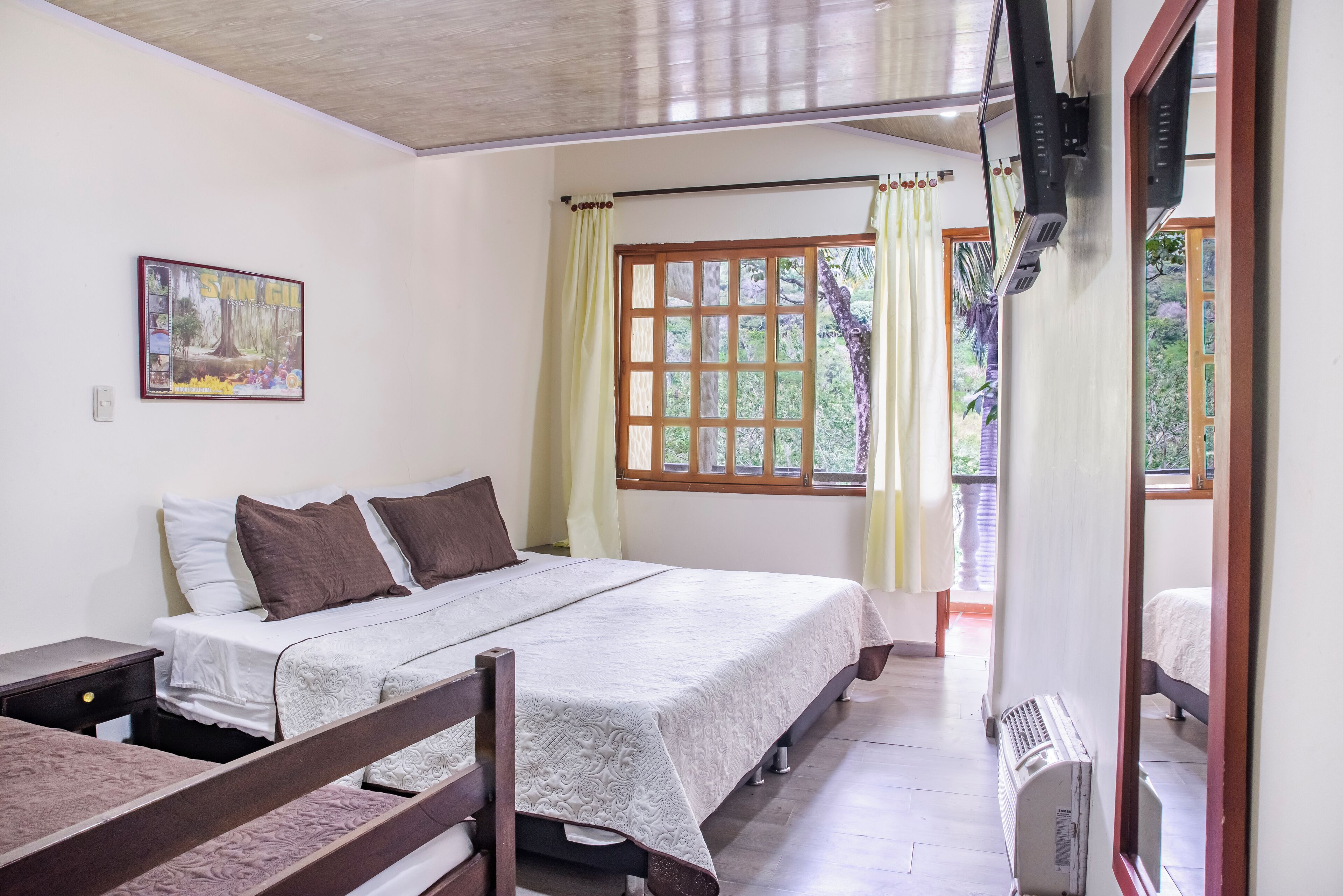 Quarto com cama, mesas de cabeceira, janela com cortinas, televisão na parede e radiador.