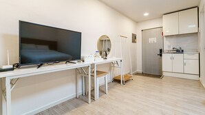 Quarto moderno e minimalista com uma televisão de tela plana grande, uma mesinha com espelho, uma cozinha compacta e uma porta que dá para o banheiro.