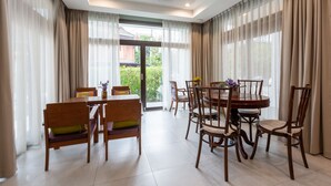 Uma área de refeições com móveis de madeira, uma mesa redonda e cadeiras. Há cortinas nas janelas e uma luminária no teto.