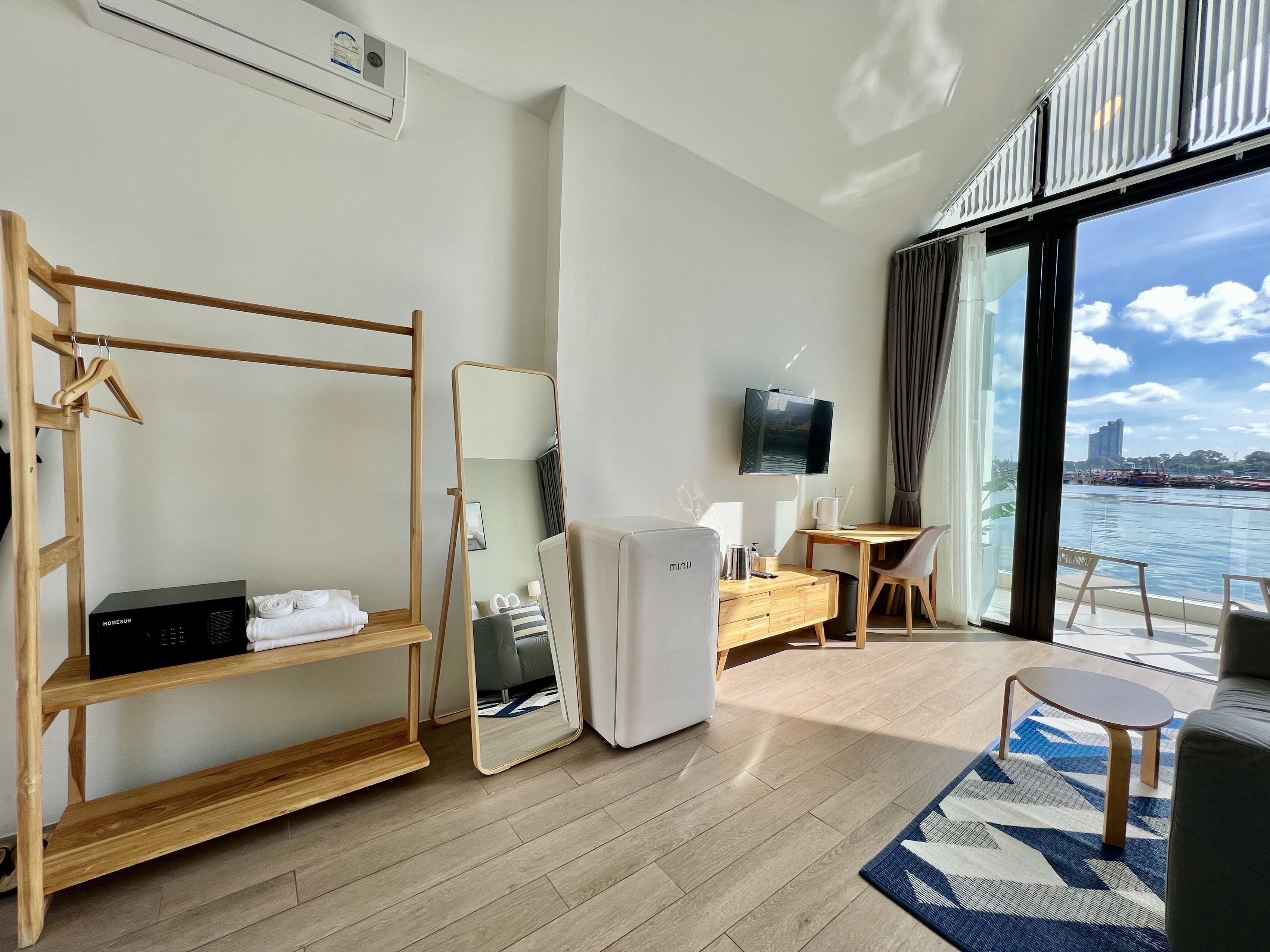 Quarto de hotel moderno com uma cama beliche de madeira, uma escrivaninha com cadeira, um pequeno frigorífico, uma TV e uma grande janela com vista para a água.