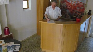 Uma área de recepção com balcão de madeira, um quadro na parede e um vaso vermelho no chão.