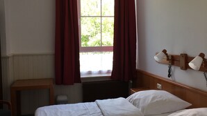 Quarto de hotel com duas camas, cabeceira de madeira, mesa de cabeceira, janela com cortinas vermelhas e uma luminária pendente.