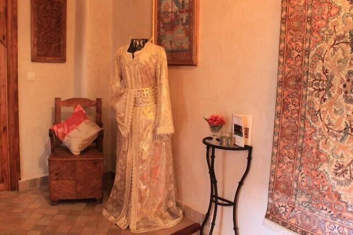 Um quarto com um manequim vestindo um vestido longo e transparente com padrão, uma pequena mesa com um vaso e uma revista, uma cadeira de vime com almofada vermelha e um tapete grande e ornamentado.