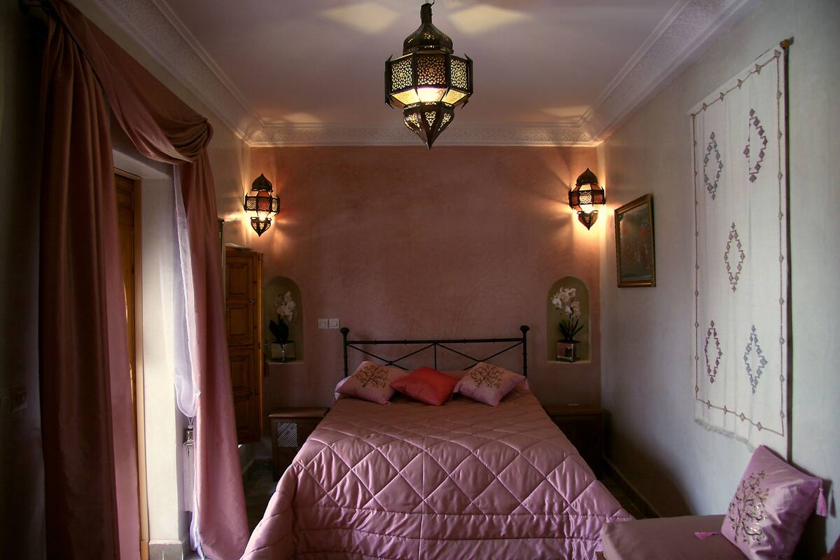 Quarto com cama, duas luminárias de parede, um ornamento pendurado na parede e janela com cortinas.