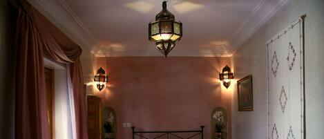 Quarto com cama, duas luminárias de parede, um ornamento pendurado na parede e janela com cortinas.