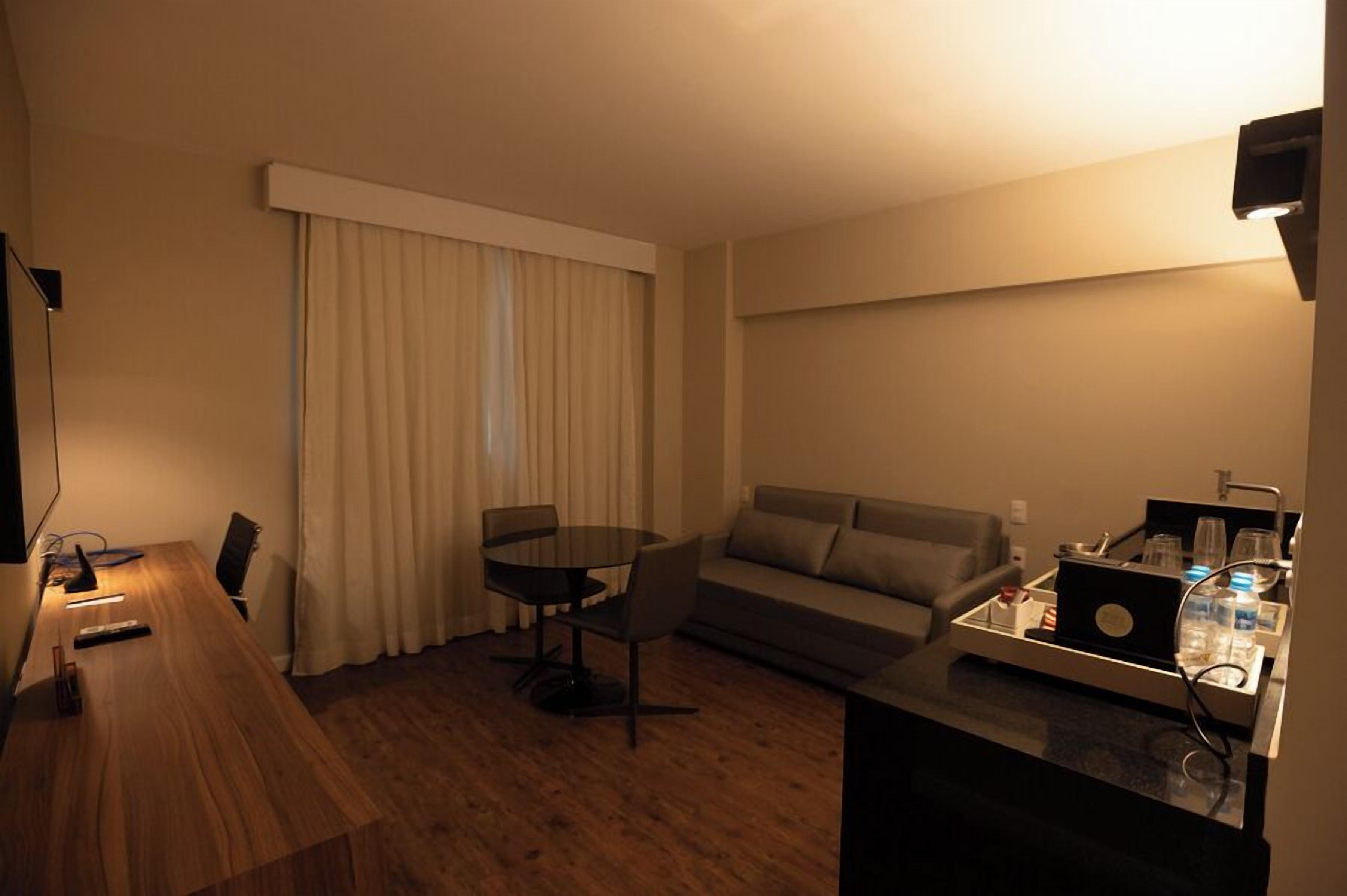 Quarto de hotel compacto com uma mesa de jantar de madeira, um sofá, uma pequena mesa redonda e um minibar.