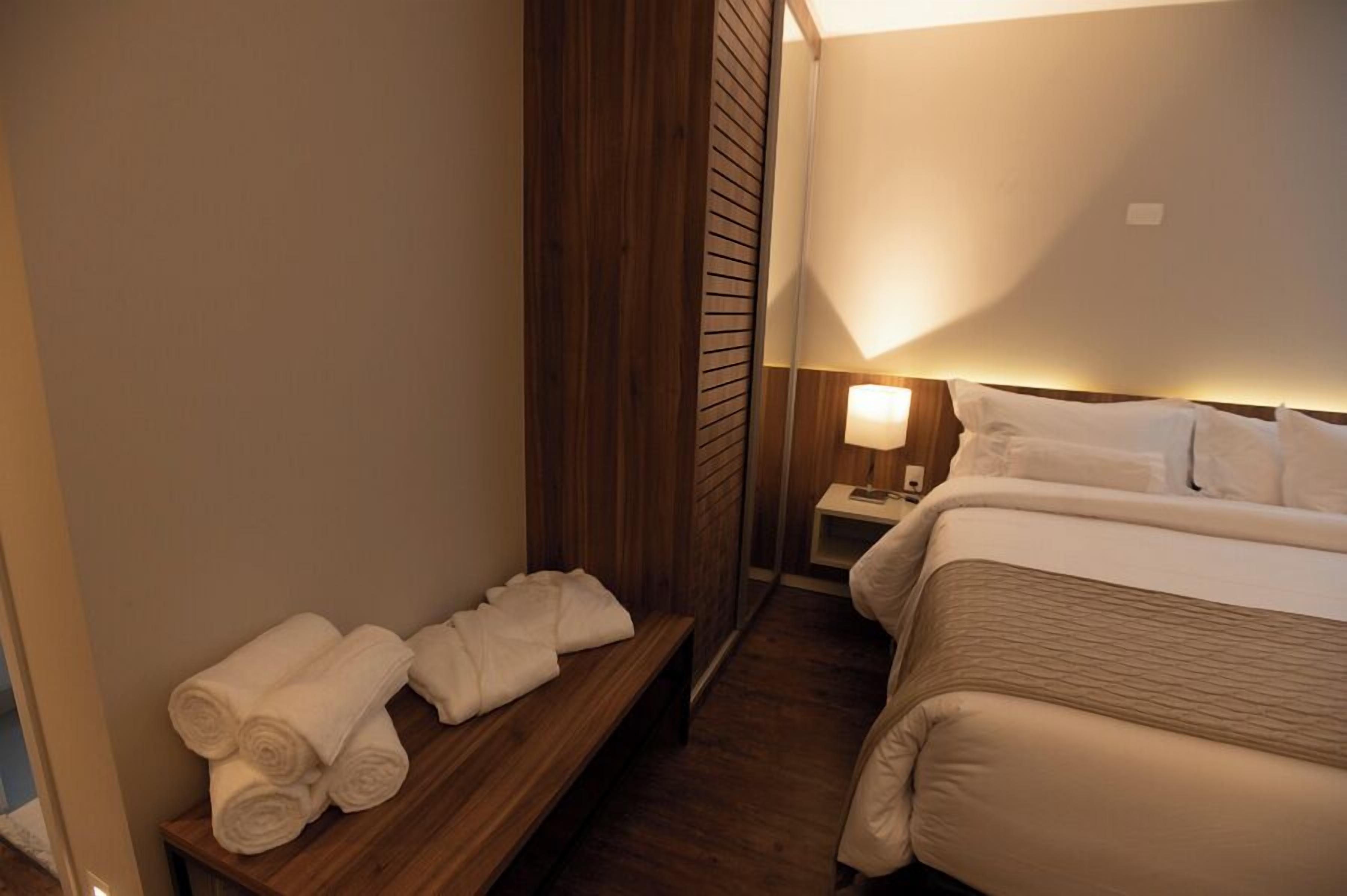Quarto de hotel com cama, mesa de cabeceira, abajur e um banco com toalhas.