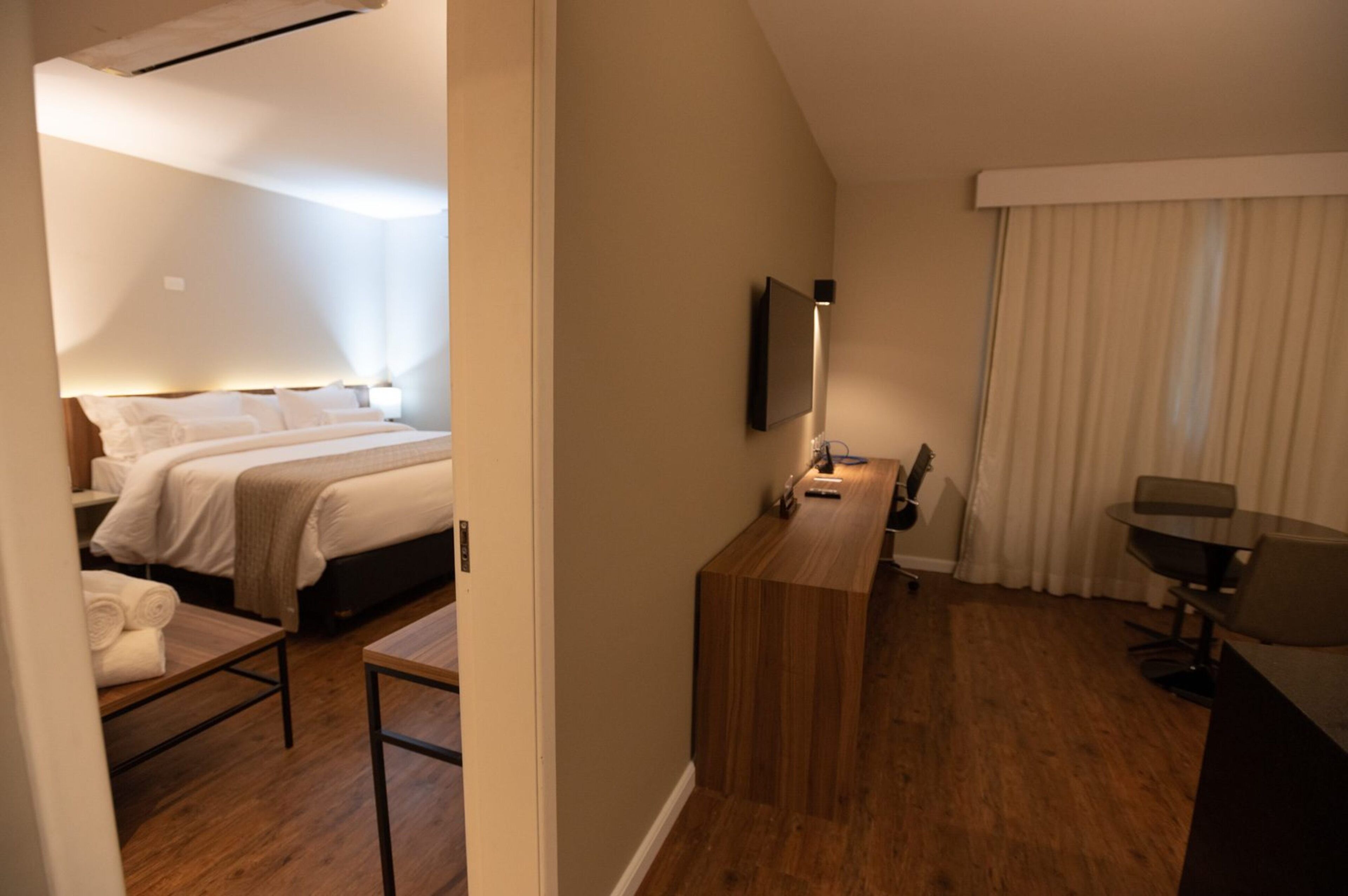 Quarto de hotel com cama, escrivaninha, cadeira, televisão e janela com cortinas.