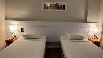 Quarto de hotel com duas camas de solteiro, cada uma com uma mesa de cabeceira e uma luminária. Há uma pequena prateleira com objetos decorativos acima das camas.