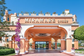A entrada do hotel Miramare Queen, com um arco imponente, uma placa sobre a entrada e um jardim bem cuidado.