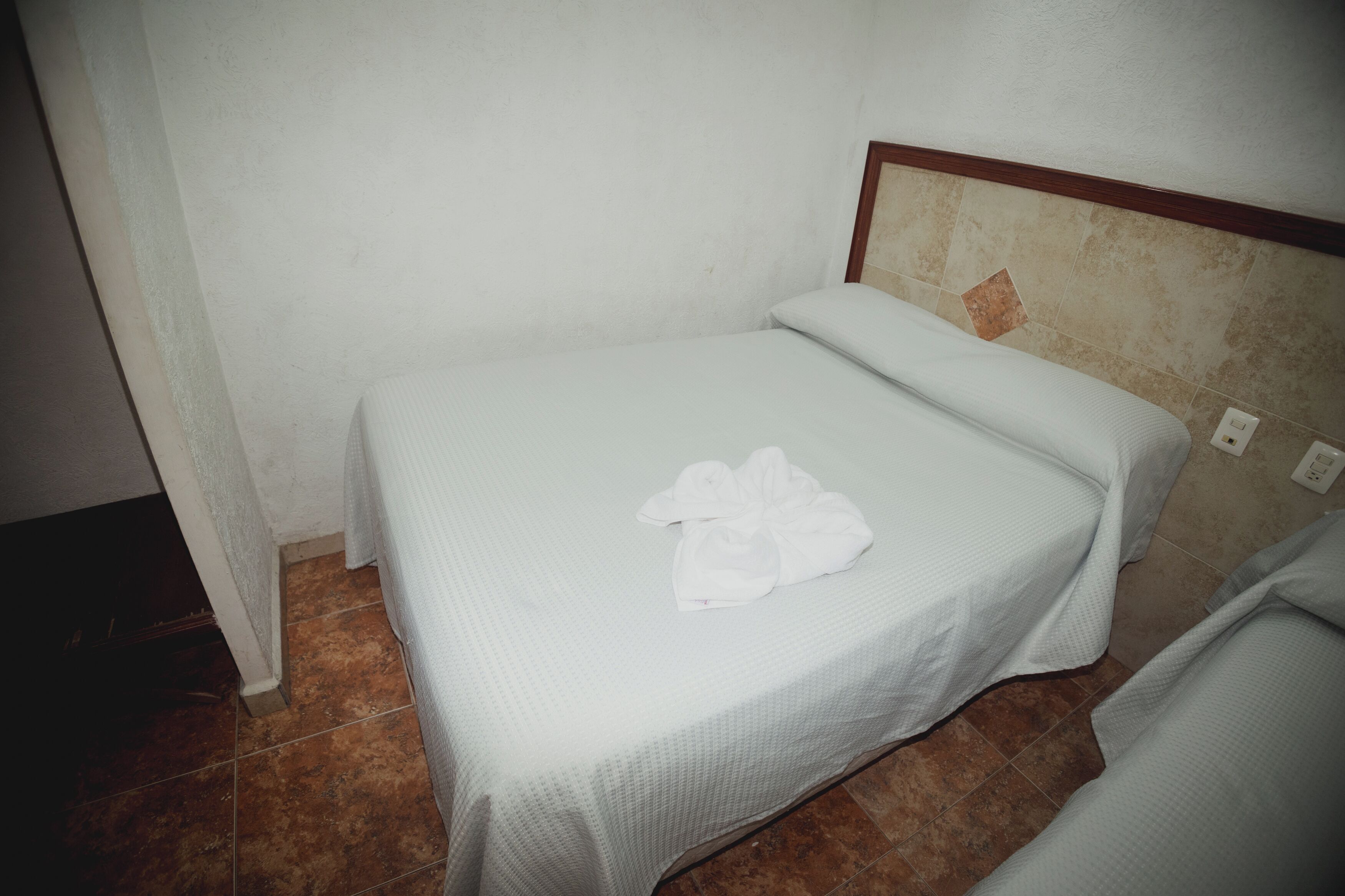 Quarto pequeno com uma cama de solteiro, parede branca sem adornos e cabeceira de madeira simples.