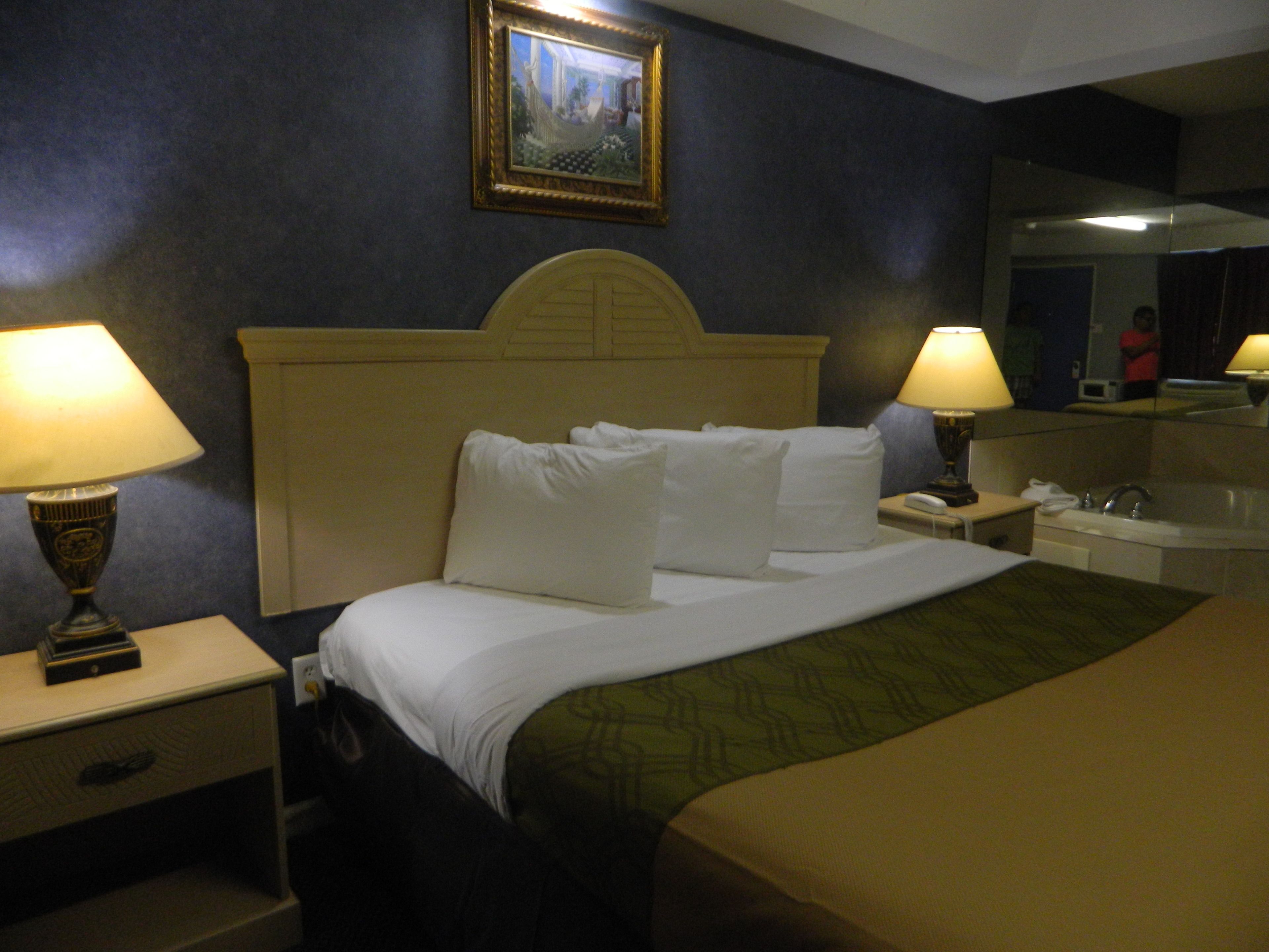 Quarto de hotel com cama, duas mesas de cabeceira com abajures, um criado-mudo, espelho e pia.