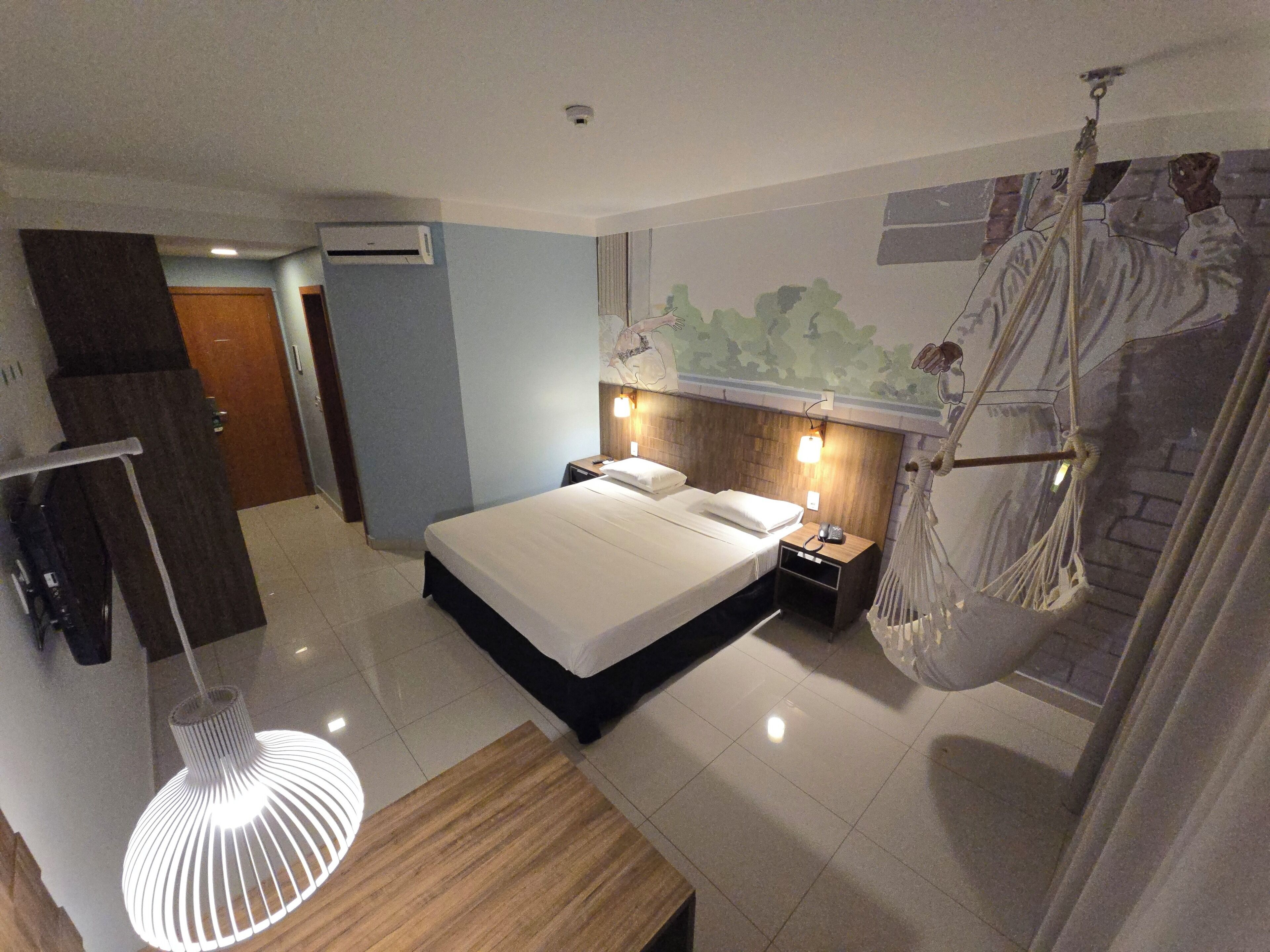 Quarto de hotel moderno com uma cama grande, mesas de cabeceira, uma cadeira suspensa e um mural decorativo na parede.