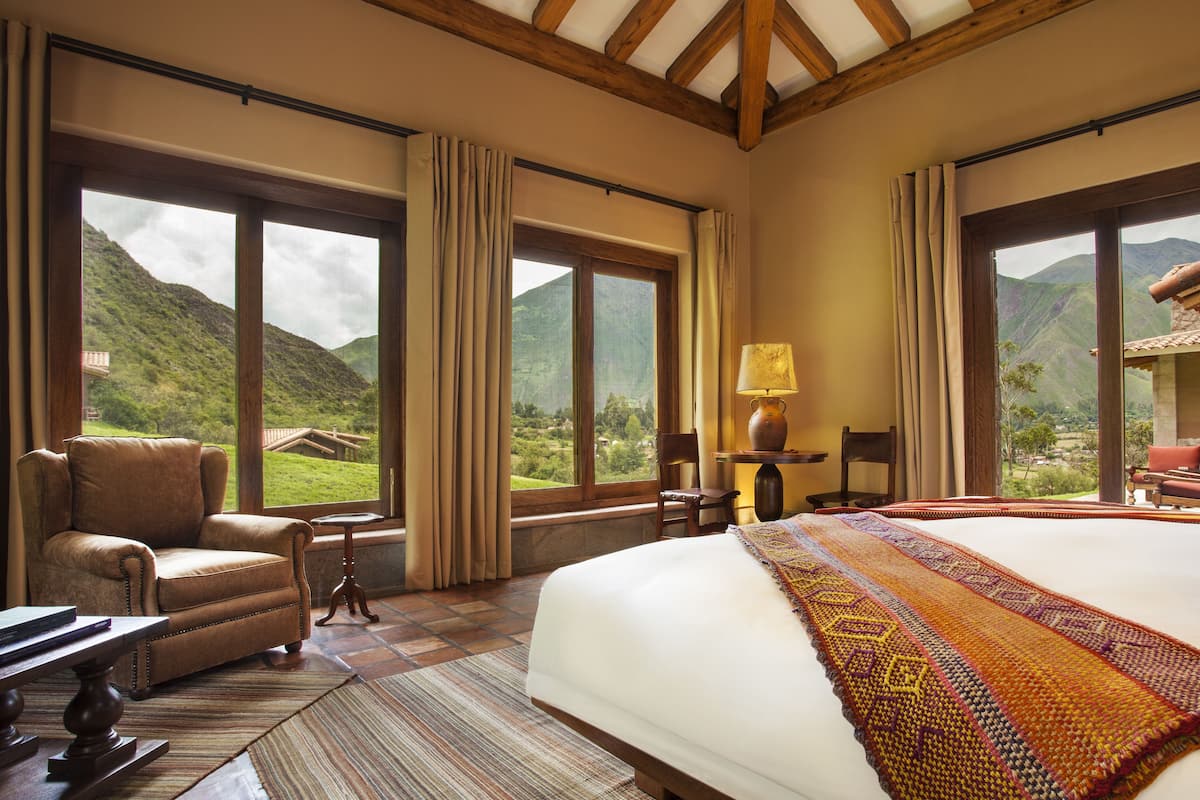 Um quarto espaçoso com uma cama grande, um espaço de estar com uma poltrona e mesa, e vista para montanhas através de grandes janelas.