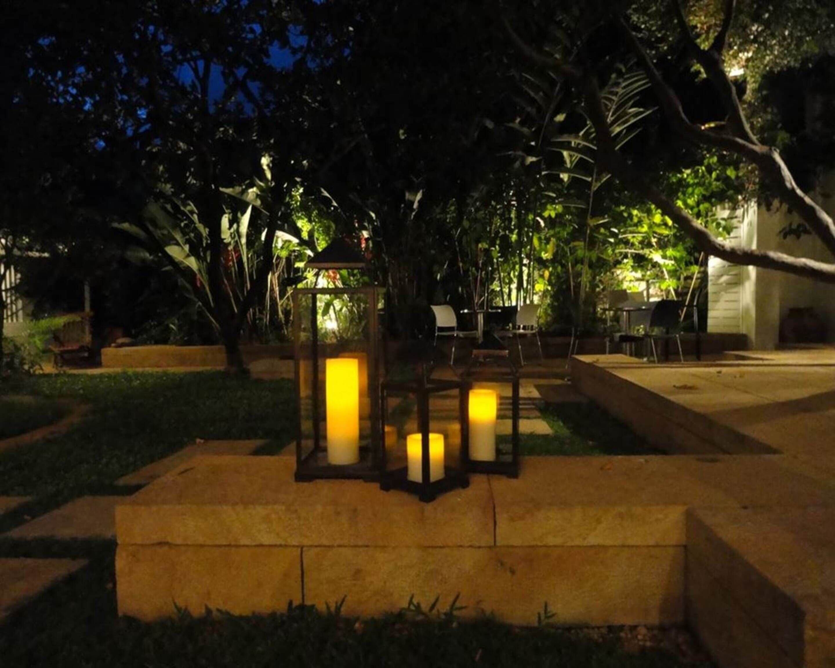 Uma área de estar ao ar livre com mesas e cadeiras, cercada por vegetação e iluminada por lanternas.