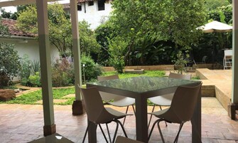 Um pátio externo com mesa de vidro e quatro cadeiras, rodeado por vegetação exuberante e um edifício branco ao fundo.