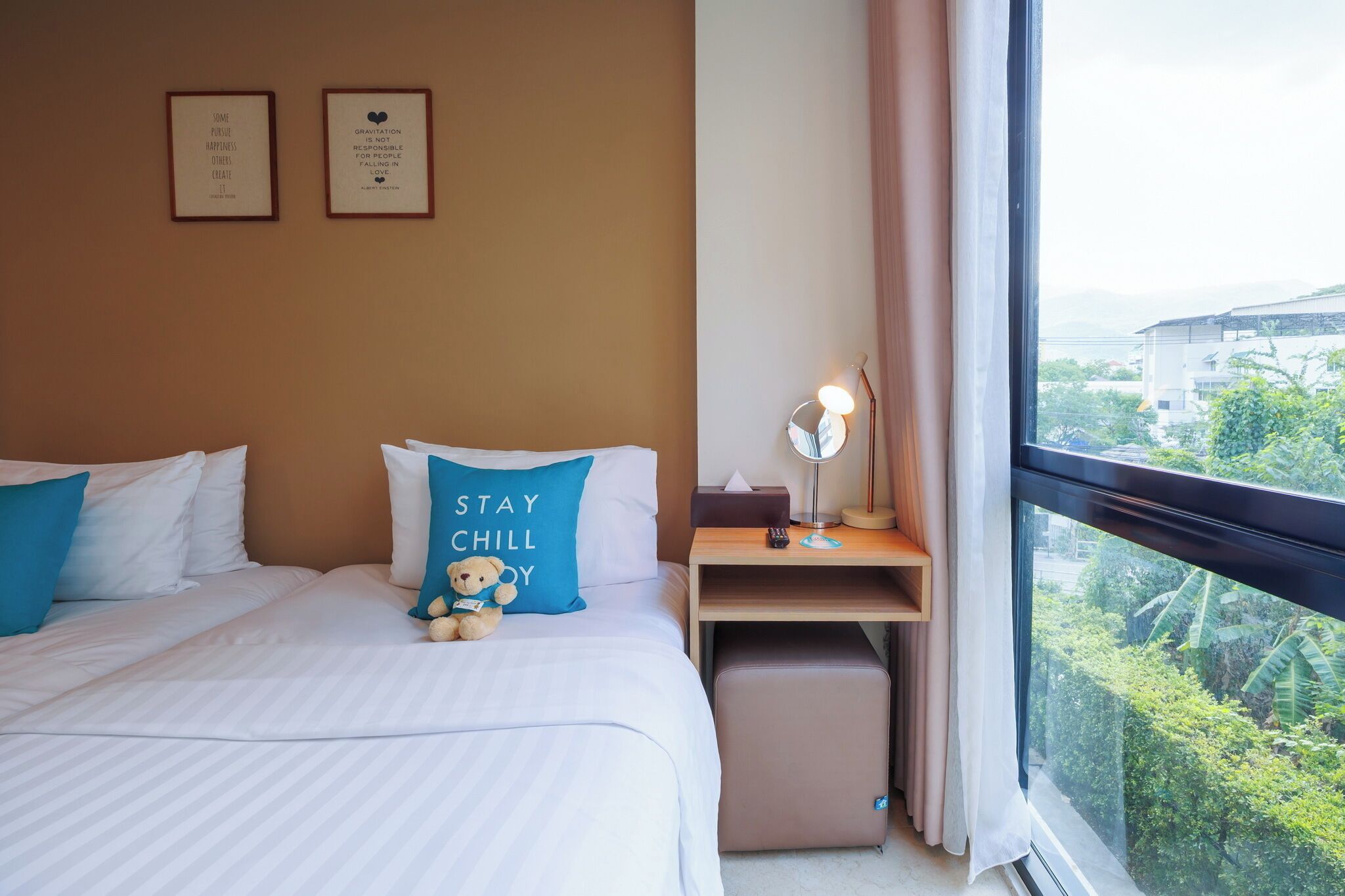 Quarto de hotel com uma cama, mesa de cabeceira com abajur, um banquinho e janela com vista para o verde.