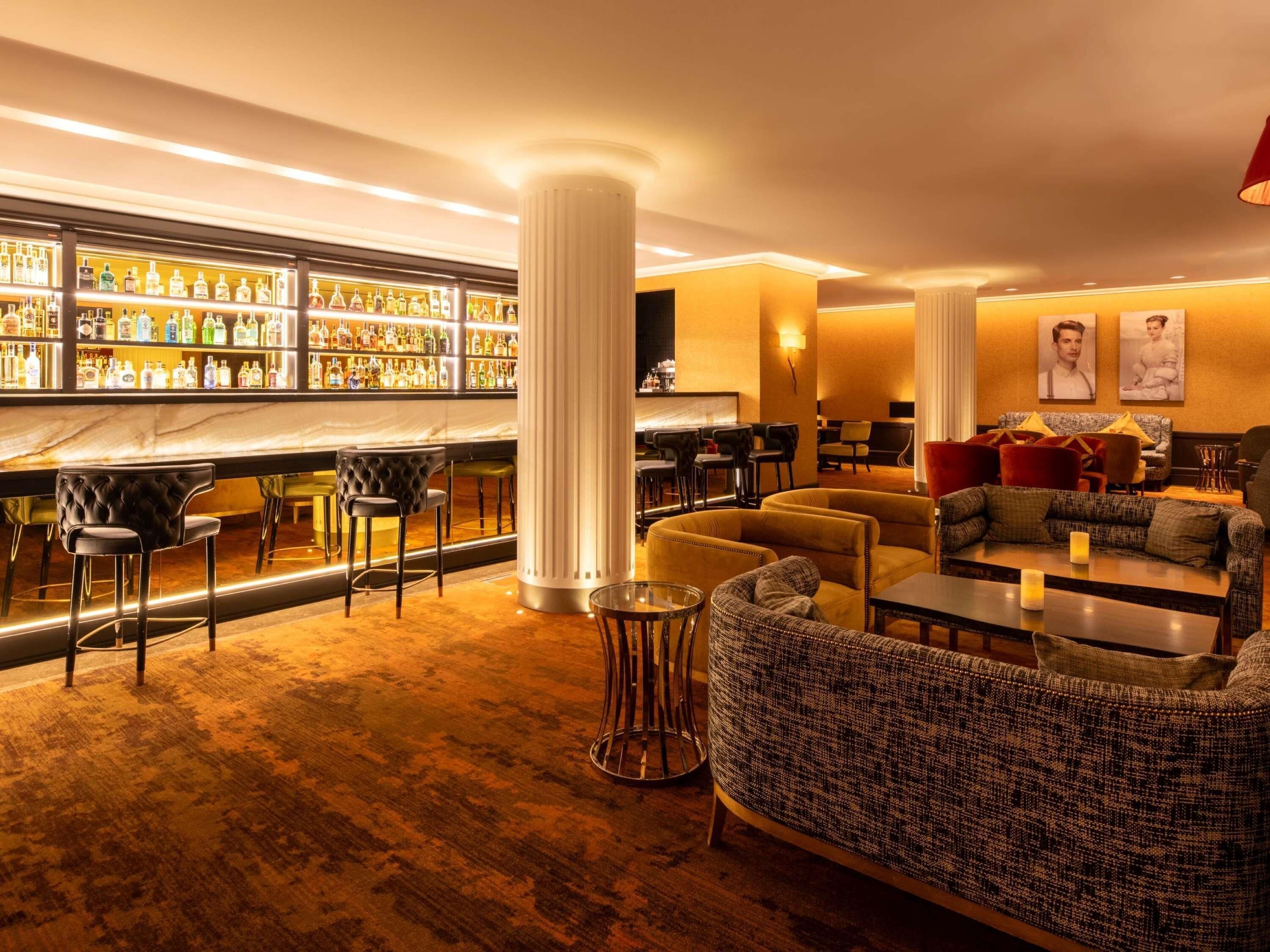 Uma área de bar com bancada de mármore e banquetas, uma sala de estar com sofás e poltronas, e uma parede com quadros emoldurados.