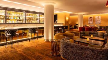 Uma área de bar com bancada de mármore e banquetas, uma sala de estar com sofás e poltronas, e uma parede com quadros emoldurados.