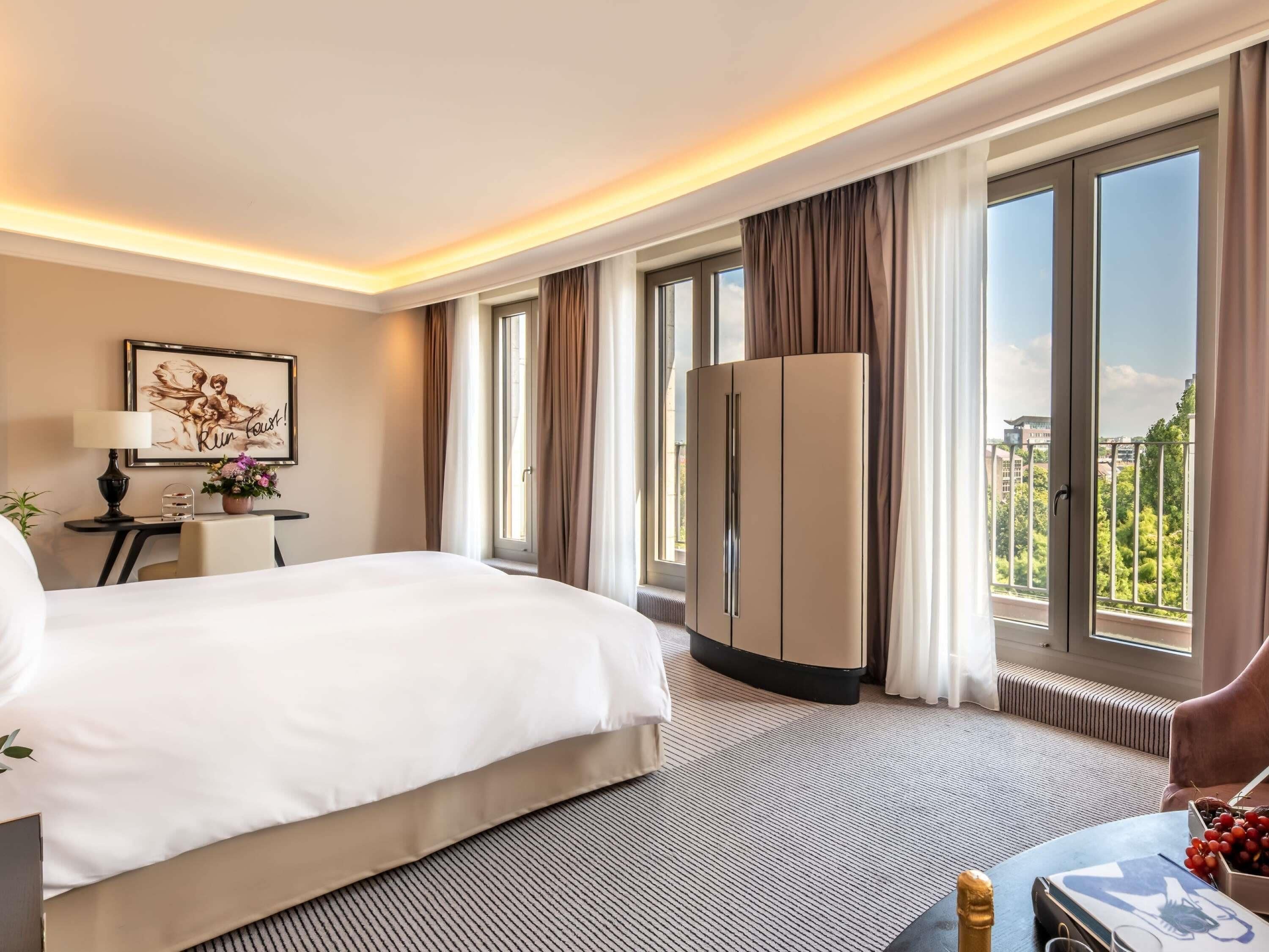 Quarto de hotel com uma cama grande, uma escrivaninha com luminária, uma planta em vaso e vista da cidade através de amplas janelas.