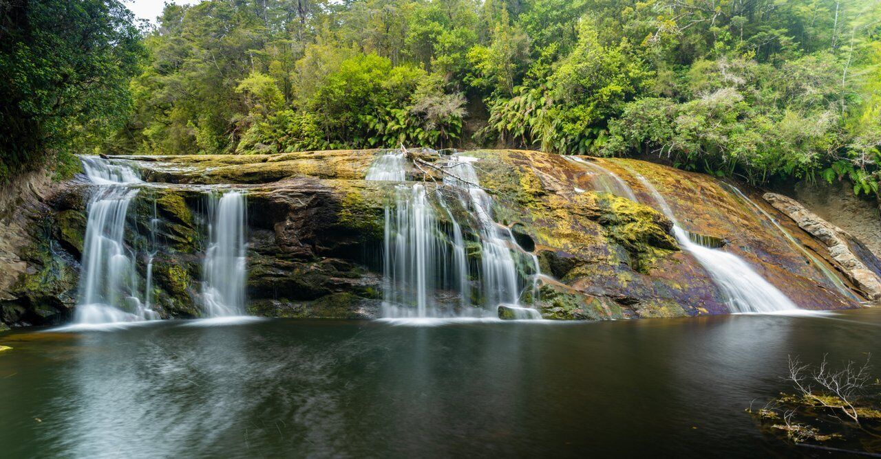 Uma cascata desce sobre um afloramento rochoso, caindo em um lago tranquilo cercado por vegetação densa e verdejante.