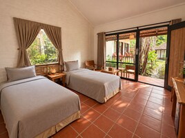Quarto de hotel com duas camas, janela com cortinas, porta de vidro deslizante para área externa com cadeiras e mesas, e piso de azulejos.