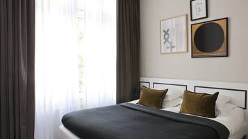 Uma cama bem arrumada com um cobre-leito escuro e duas almofadas, uma mesinha de cabeceira com abajur, e uma janela com cortinas finas permitindo a entrada de luz natural.