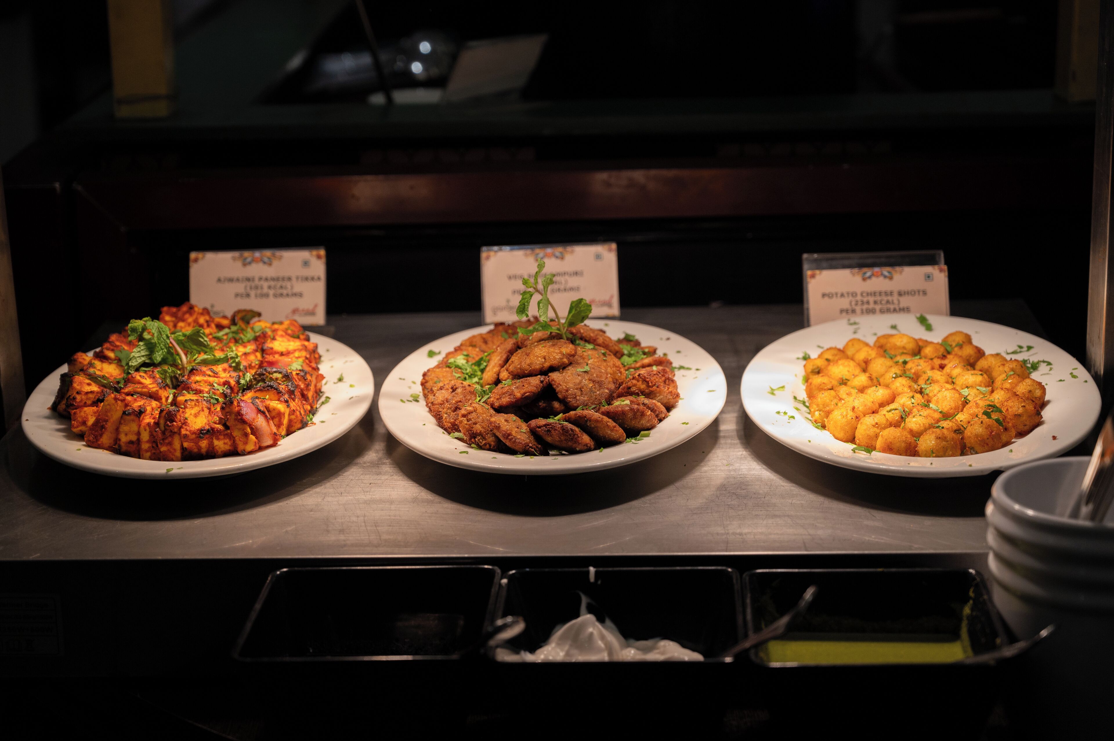 Buffet com três pratos de comida: uma pizza, frango frito e mini porções de batata com queijo.