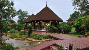 Um gazebo com telhado de azulejos, rodeado por um jardim com lago, deck de madeira e diversas plantas.
