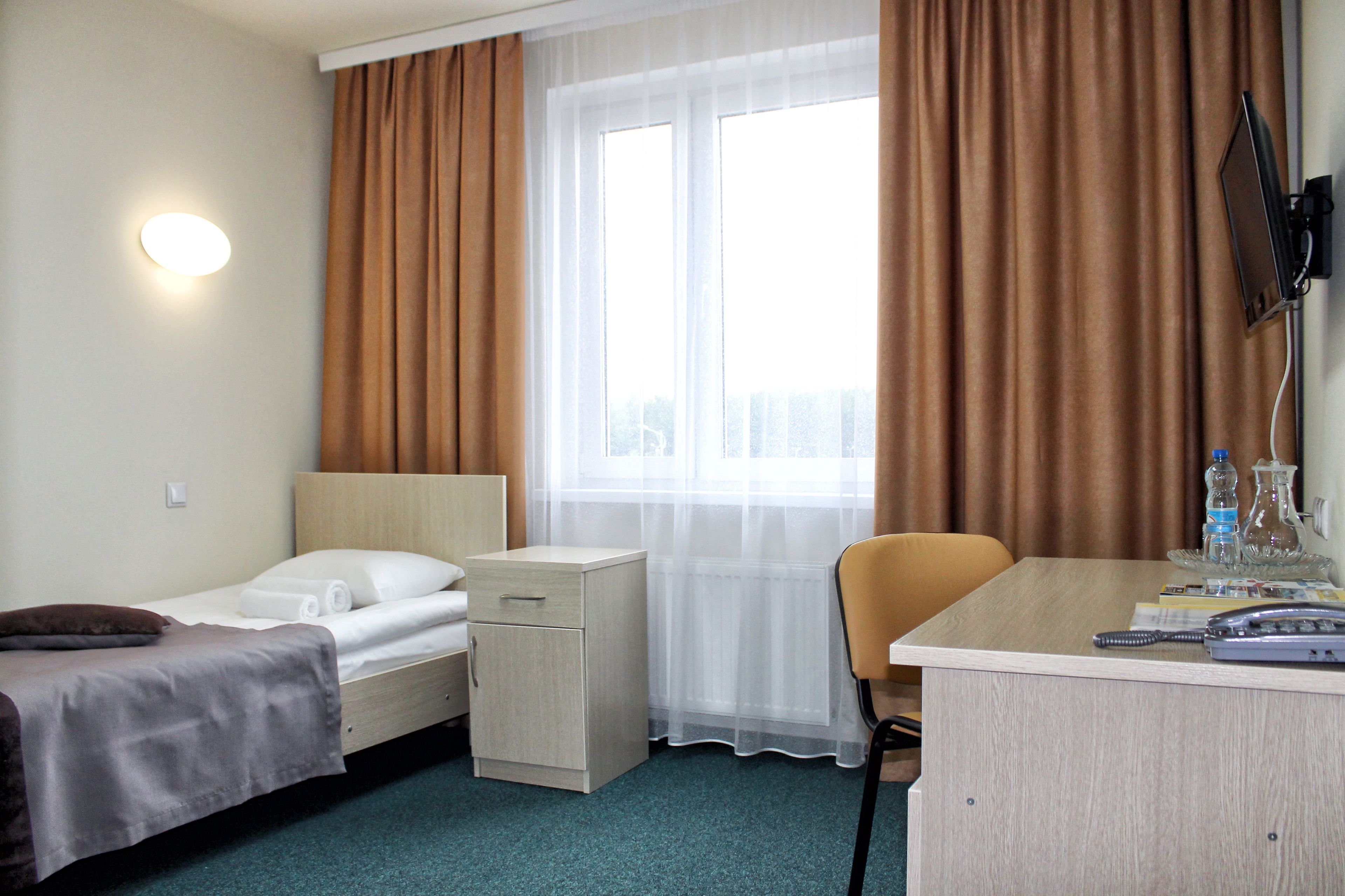 Quarto de hotel com duas camas de solteiro, uma escrivaninha com cadeira, televisão presa na parede e uma janela grande com cortinas.