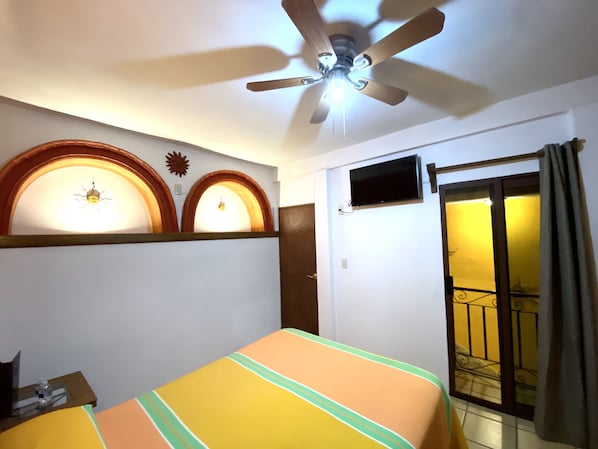 Quarto de hotel com cama, ventoinha no teto, janelas em arco e televisão.