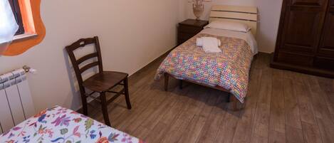 Quarto com teto de madeira, cama com colcha florida colorida, uma cadeira, guarda-roupa de madeira e um quadro na parede.