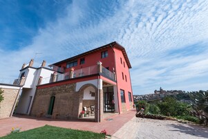 Uma villa vermelha com fachada de pedra, uma varanda e vista para uma colina com edifícios.
