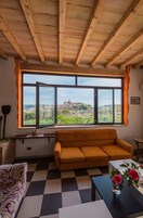 Quarto com teto de madeira, uma janela ampla com vista para uma cidade, um sofá, uma mesa de centro e uma cadeira estampada.