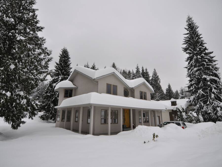 Uma casa de dois andares com telhado coberto de neve e entrada, cercada por árvores perenes.