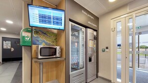 Uma área de recepção de hotel com um painel digital, máquinas de venda automática, micro-ondas e entrada por porta de vidro.
