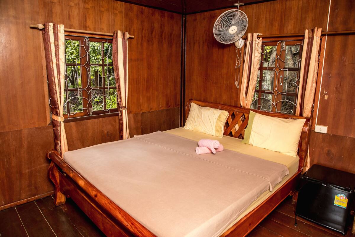 Um quarto de madeira com uma cama, um ventilador e um pequeno armário preto.