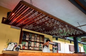 Área de bar com teto de madeira e copos pendurados. Há prateleiras com garrafas e copos atrás do balcão.