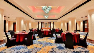 Uma sala de banquetes com várias mesas redondas, cadeiras pretas e toalhas vermelhas.